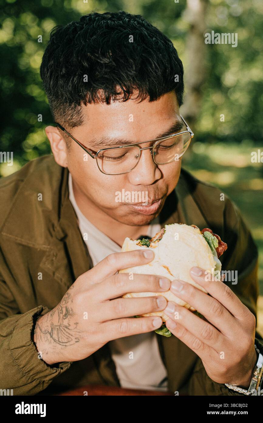 A man in a green jacket is eating a sandwich. He is wearing glasses and ...