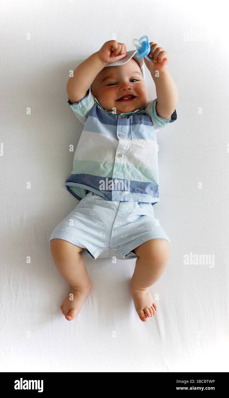 Happy 10-month-old baby boy lying on white sheet, wearing striped short ...