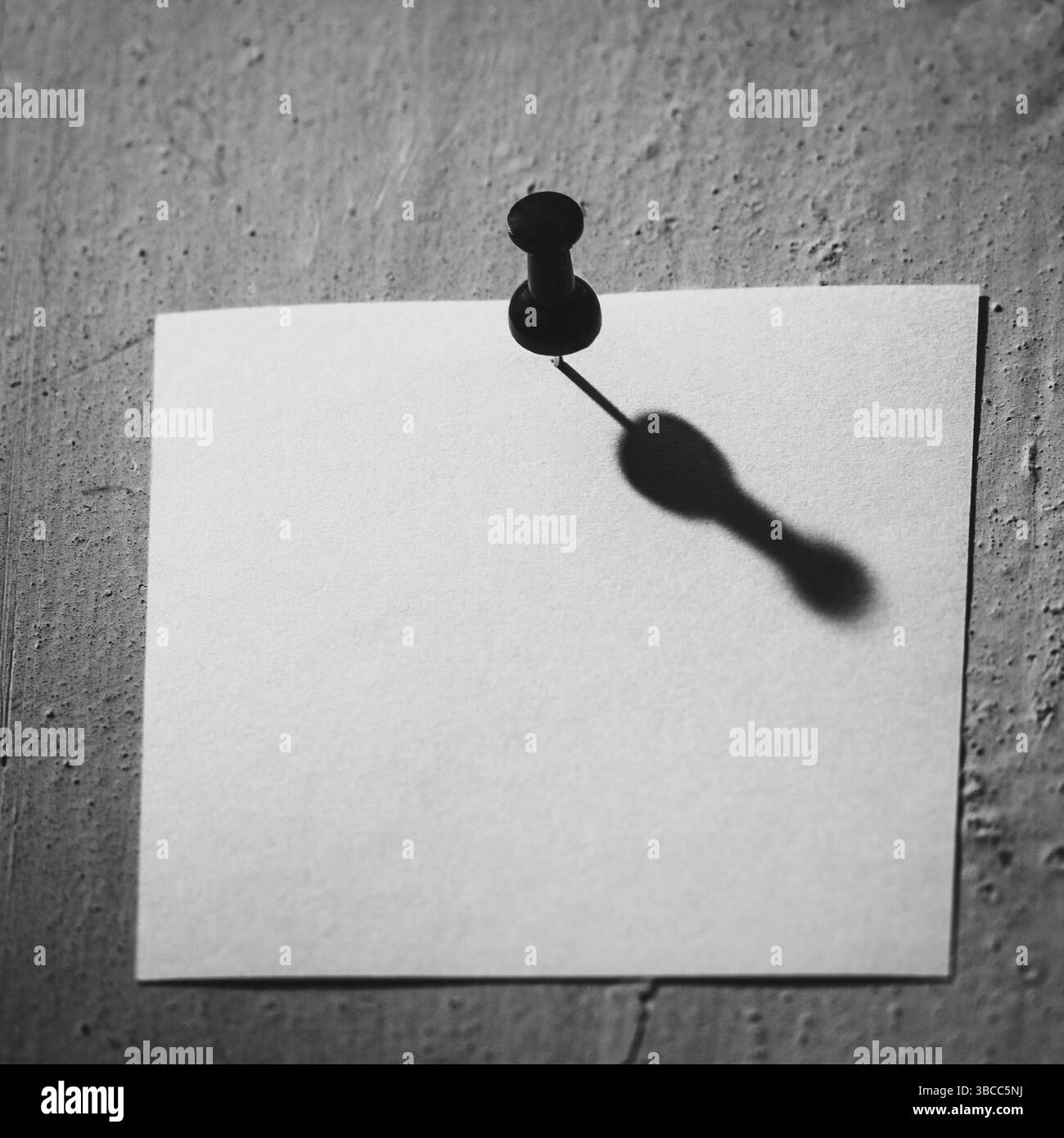 A blank piece of note paper is pinned to a textured wall, casting a ...
