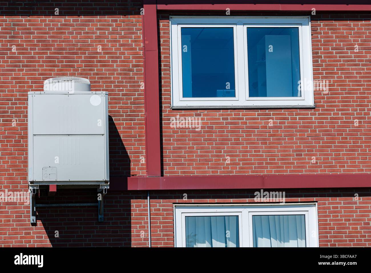 An air conditioning unit is fixed to a brick wall above two windows ...
