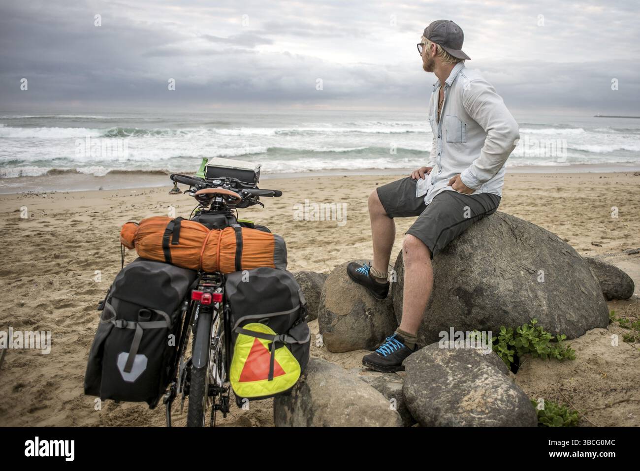 A long distance cyclists sits on a rock by the beach viewing the ocean ...