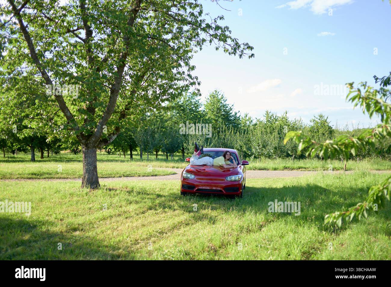 Woman poses on a red car parked under a lush tree in a sunny green ...