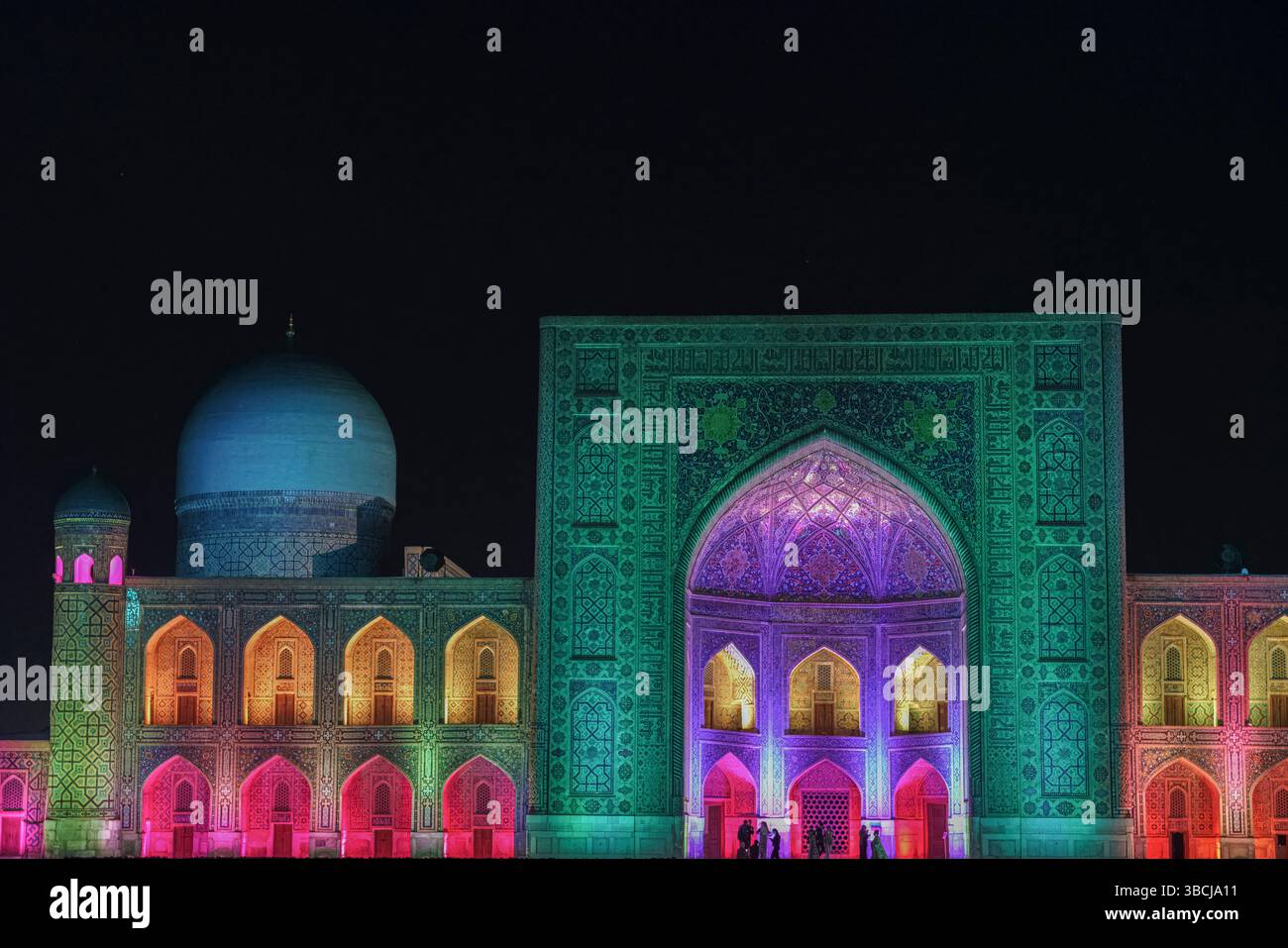 Tilya Kori Madrasa’s arched facade and blue domes glow in green and violet light at night in ...