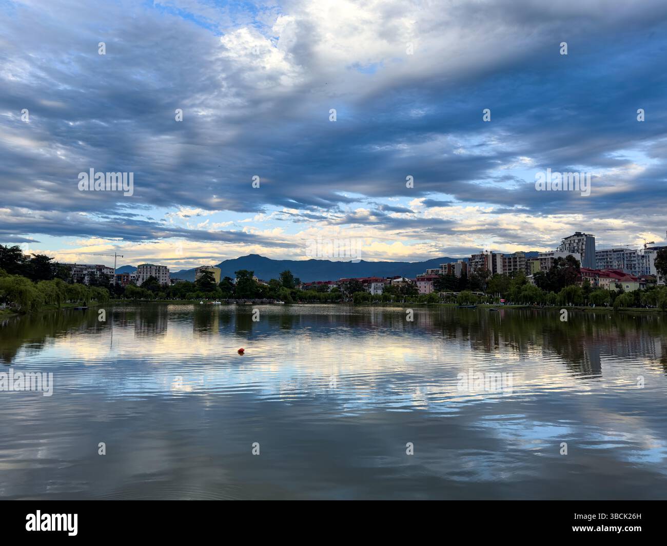 Urban lake view with modern buildings and mountain range under dramatic ...