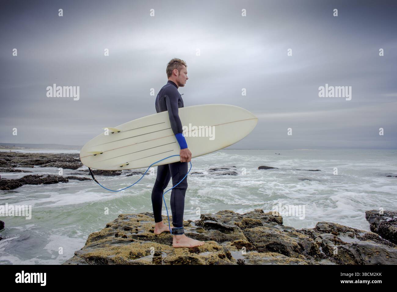 A male surfer with wetsuit and surfboard under arm stands on the rocks ...