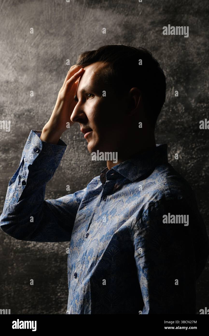 A man in a blue shirt poses with his hand on his forehead, illuminated ...