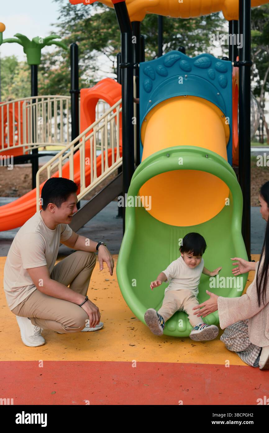 Happy parents watch and cheer as their toddler slides down a colorful ...