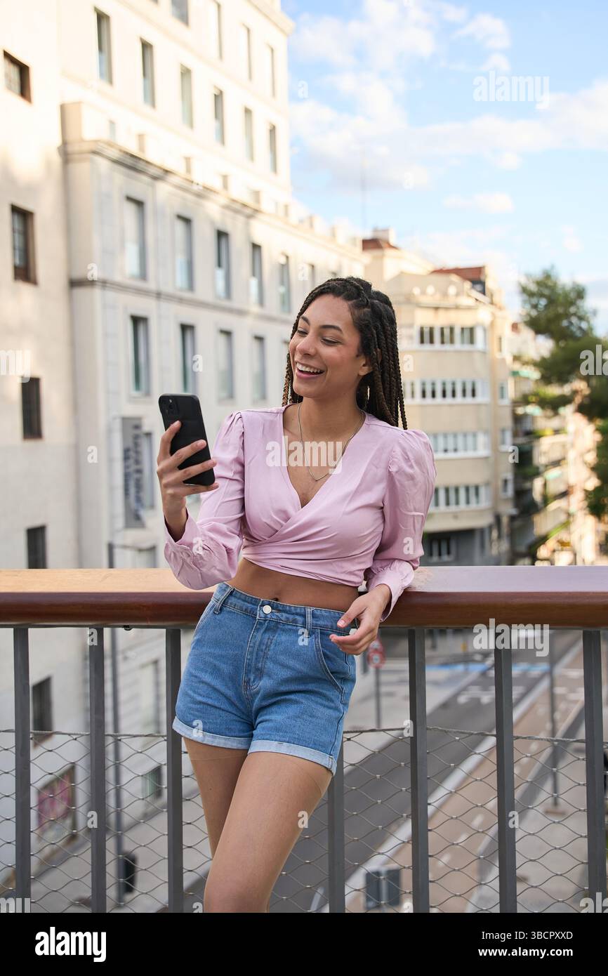 Happy young woman with braids using her smartphone, leaning on a ...