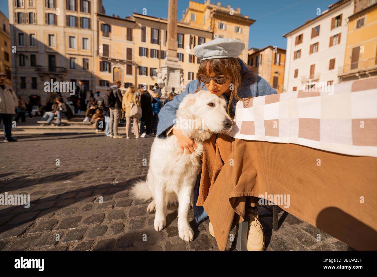A woman in a blue coat hugs her white dog beside a cafe table in a ...