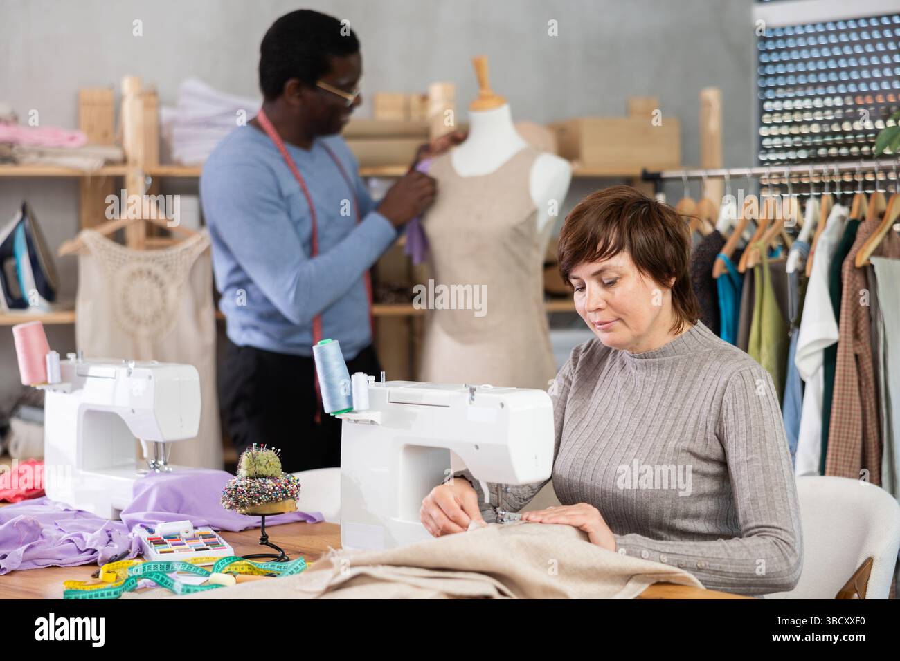 Woman tailor sews clothes, African male companion work in background ...