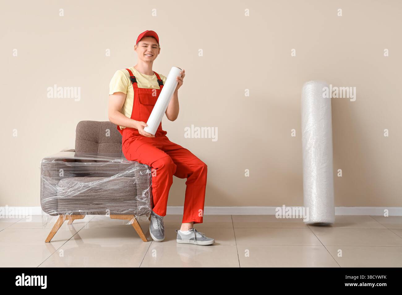 Male worker with roll of stretch wrap sitting on armchair in room Stock ...