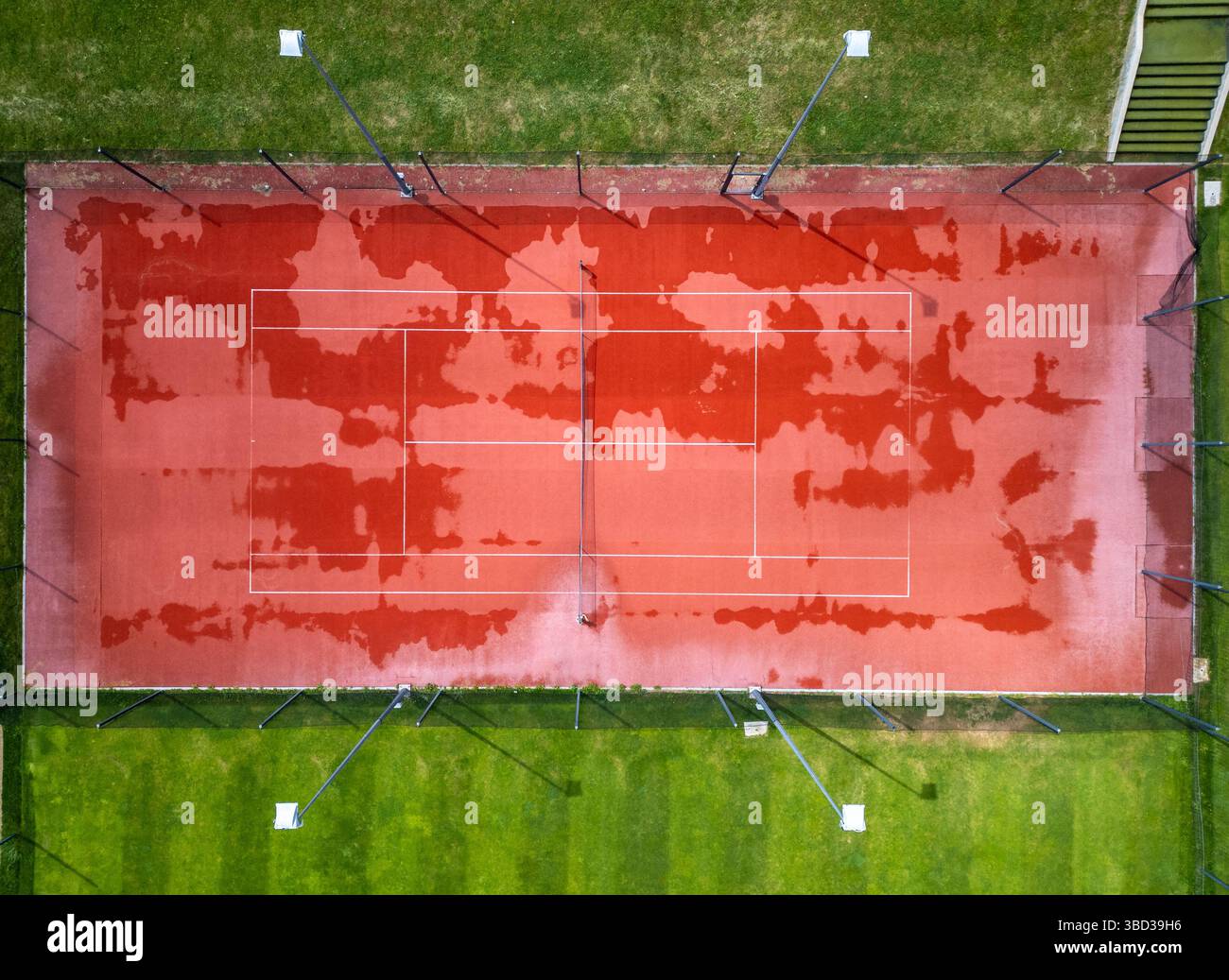 Aerial view of a red tennis court with wet patches after rain ...