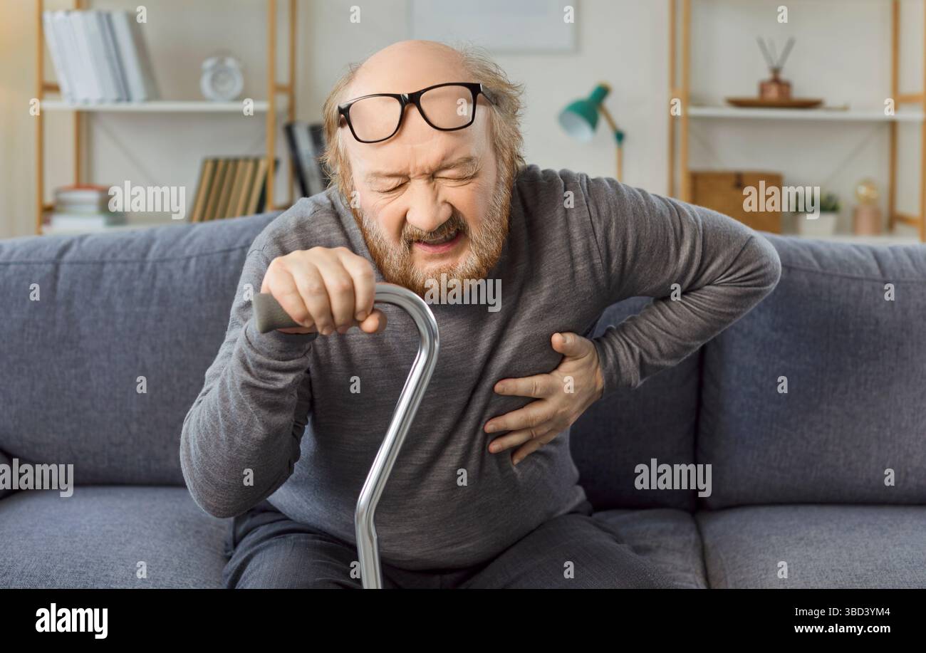 Elderly man sitting on sofa with cane, clutching chest in pain and ...