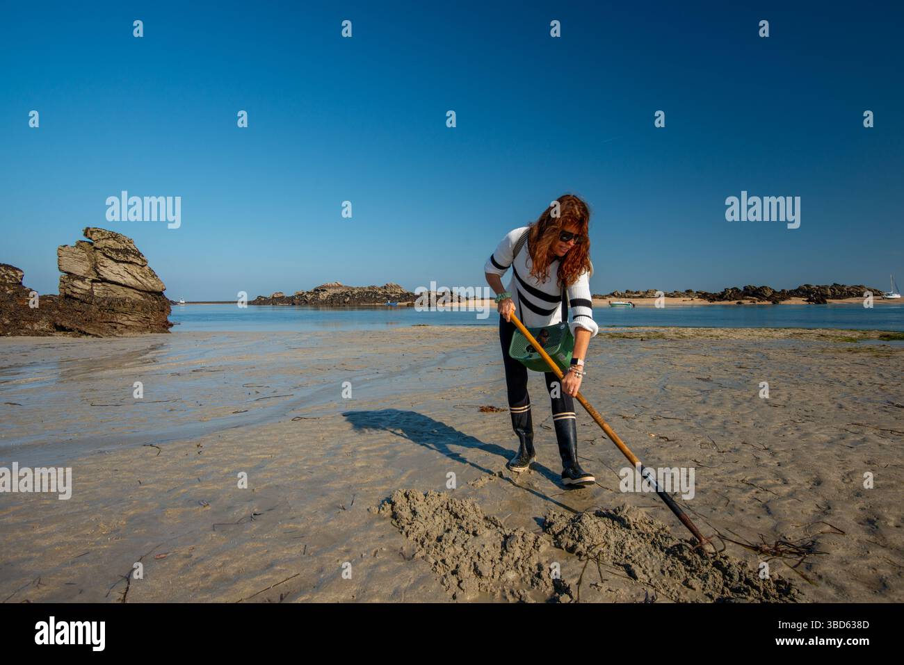 A woman dressed as a sailor with rubber boots and a hoe raking the sand ...