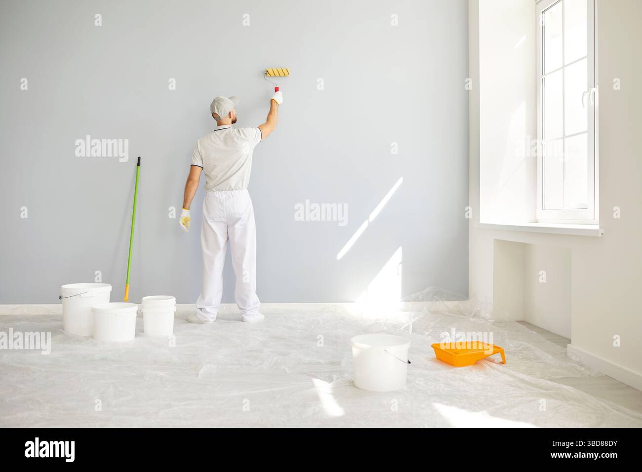 Male painter in uniform painting grey wall with roller in bright room ...
