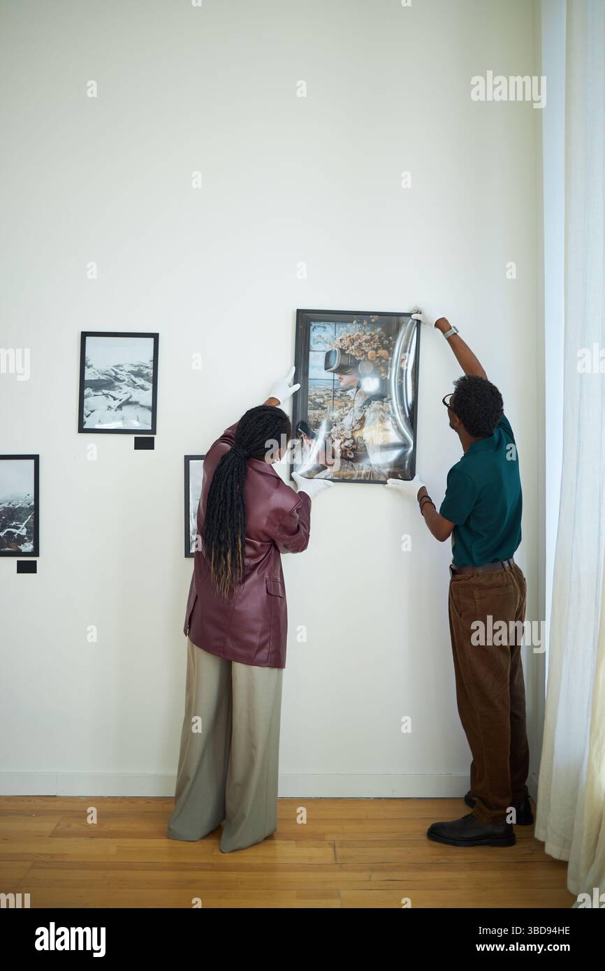 Two people rearranging framed photos on clean white gallery wall ...