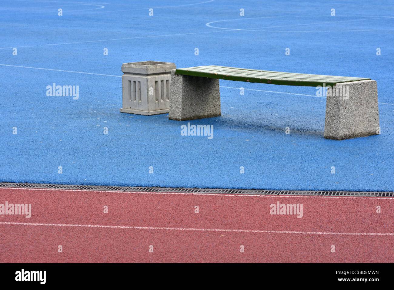 Blue sports field, red running track and empty bench with trash bin in ...