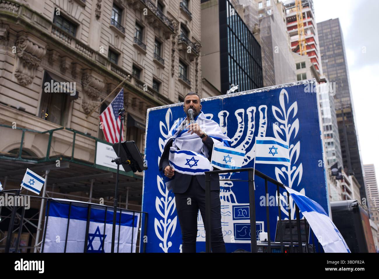 A man in a suit speaks into a microphone, draped in an Israeli flag, at ...