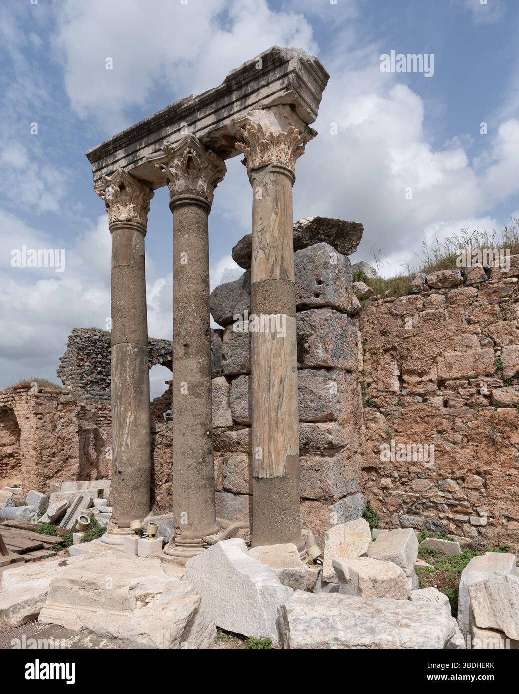 Historic ruins featuring three ancient columns with ornate capitals and stone walls at Ephesus ...