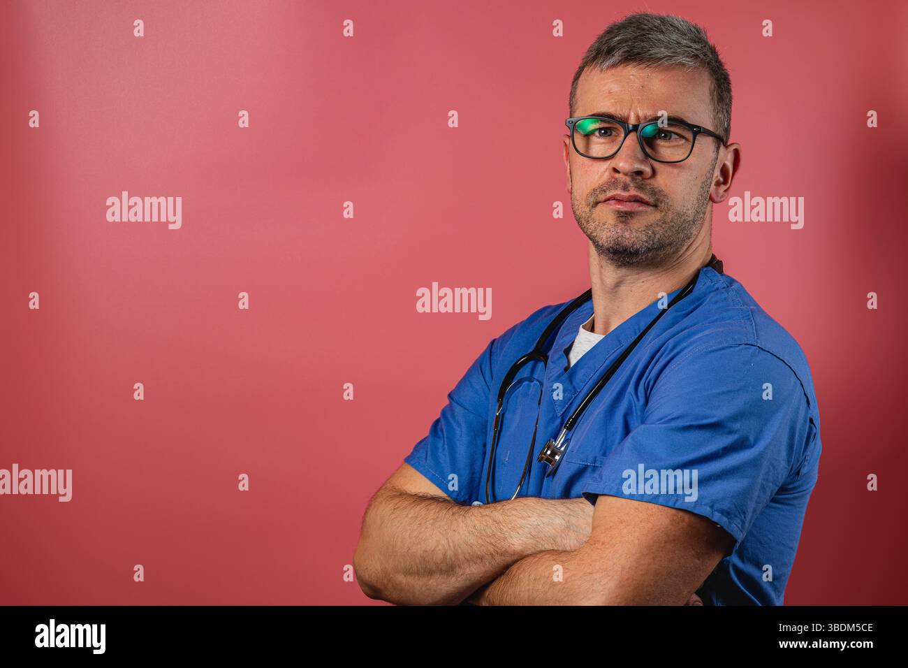 A male doctor wearing blue scrubs and glasses stands with arms crossed ...