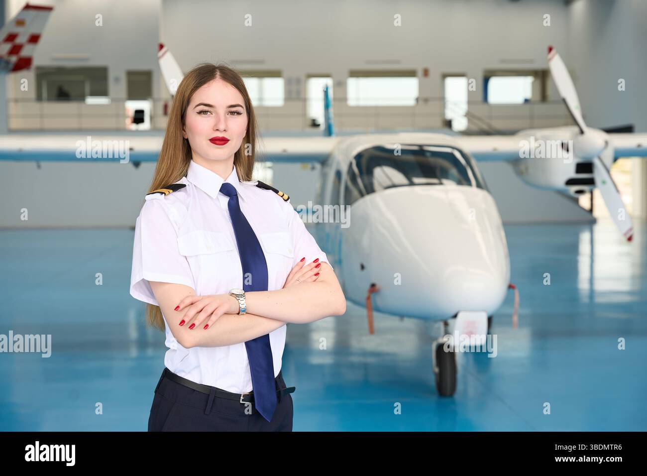 Confident female pilot posing with arms crossed in aircraft hangar ...
