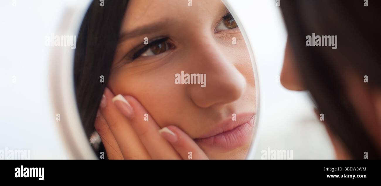 A young woman looks at her reflection in a mirror, gently touching her ...