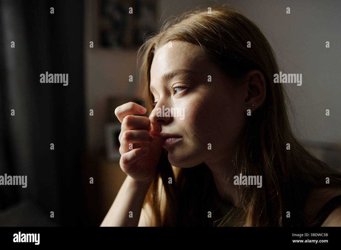 Woman sitting indoors resting her chin on hand and gazing out window ...