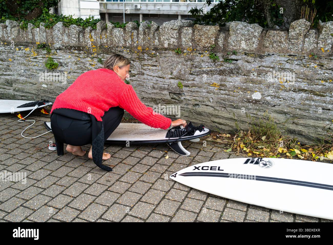 A female surfer waxng her surfboard in a car park in Newquay Towan ...