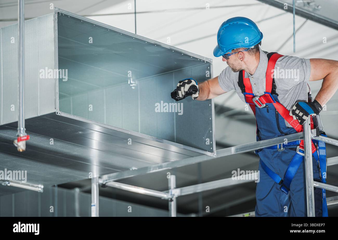 A HVAC worker in a blue helmet and gloves is securing ductwork inside a ...