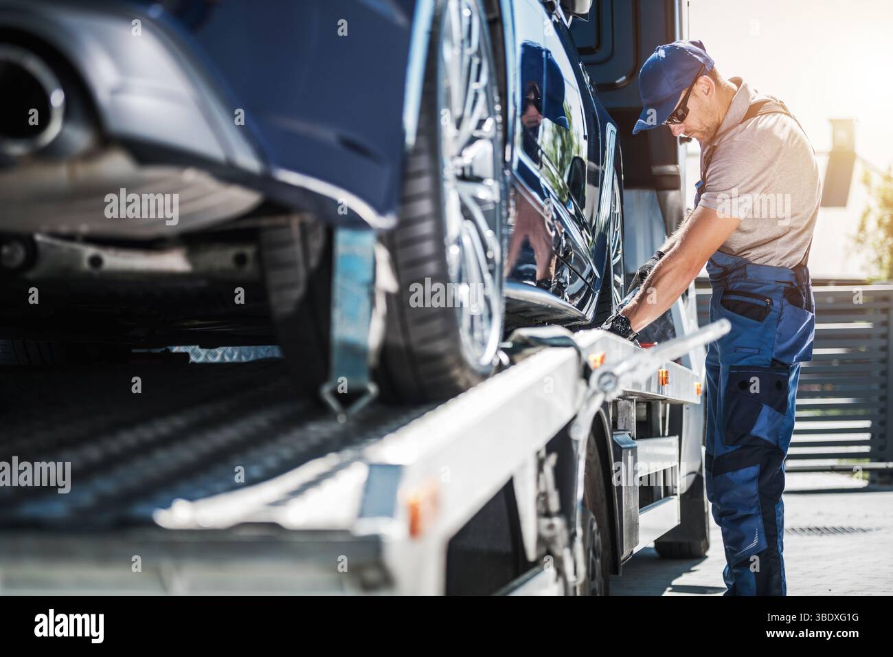 A worker secures a blue vehicle onto a transport truck in bright ...