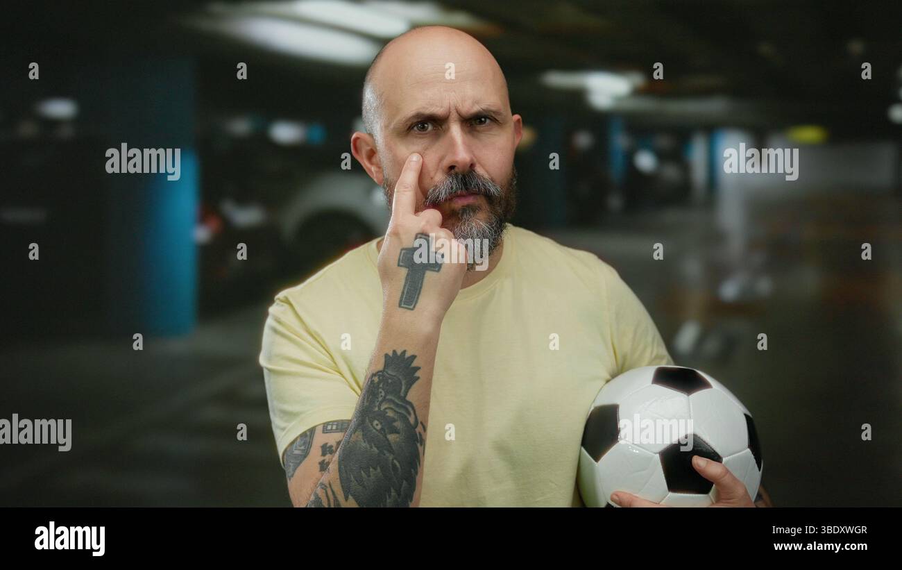Bald man with a beard holding a soccer ball in an indoor parking area ...
