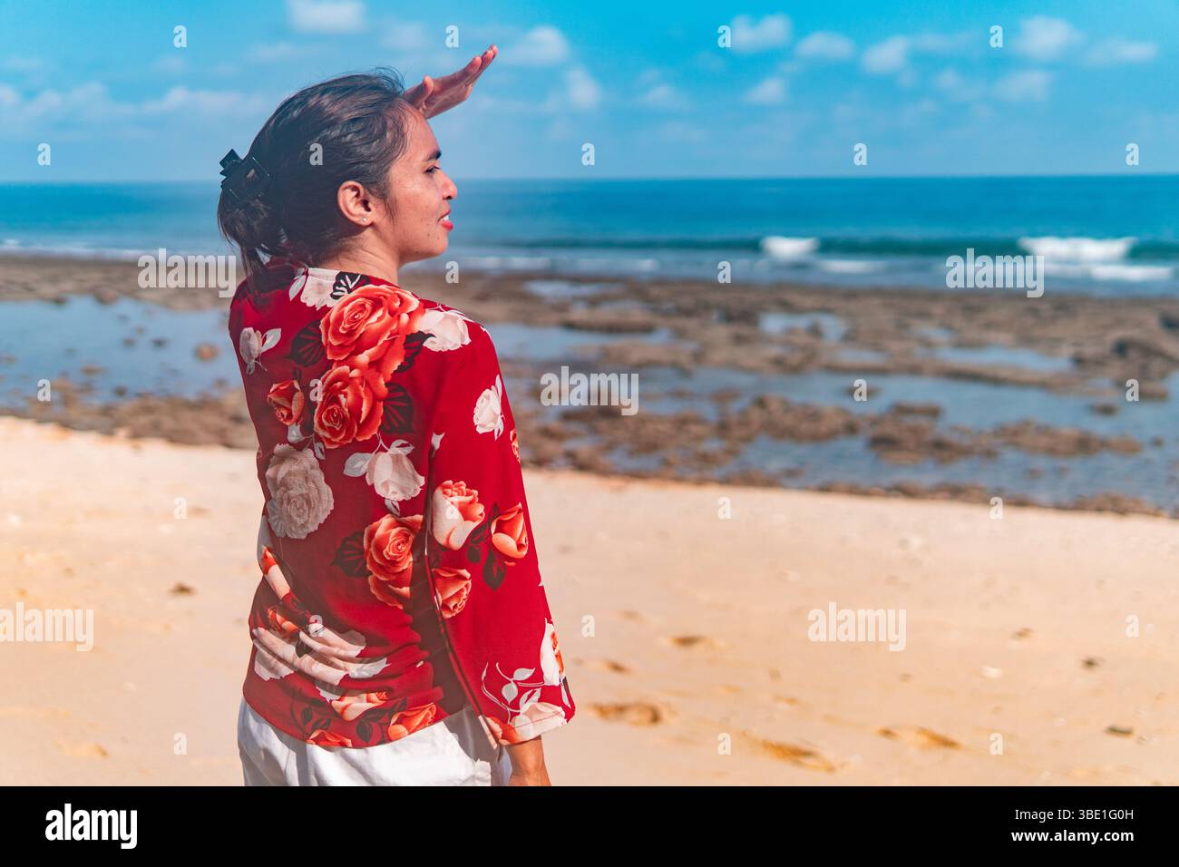 Young woman shielding her eyes from the sun while enjoying the ocean view from a beautiful beach ...