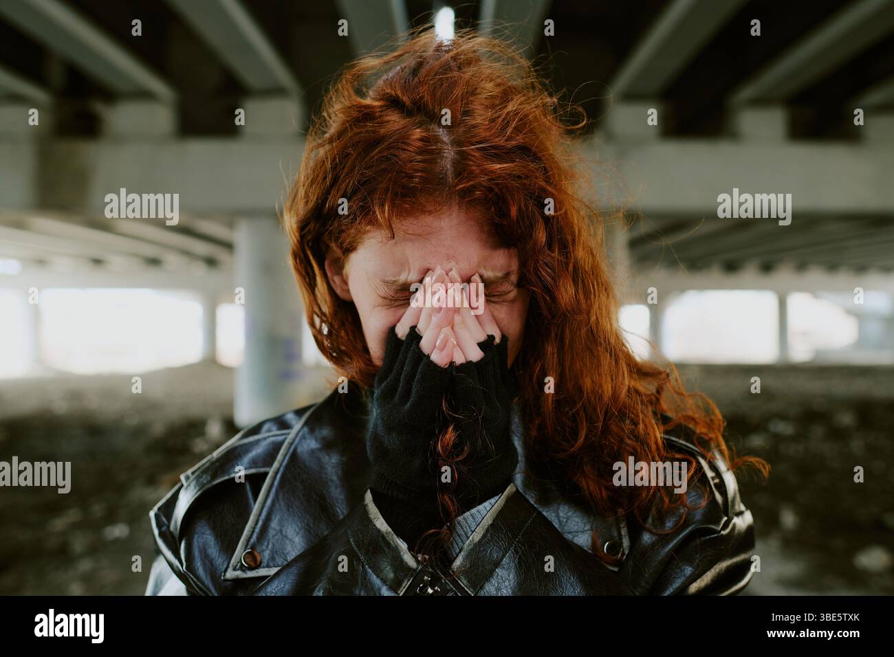 Red-haired individual sitting under bridge covering face with hands ...