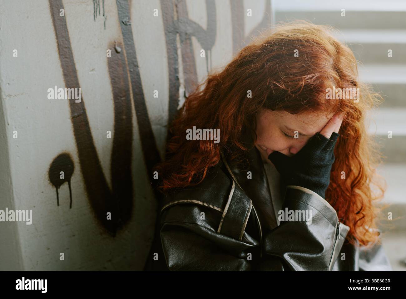 Woman with red hair in leather jacket standing against graffiti wall ...