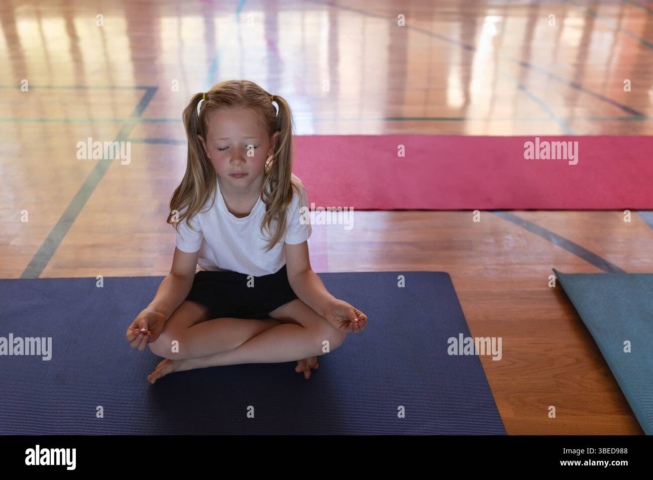 Girl sitting cross-legged on blue yoga mat on polished wooden hall ...