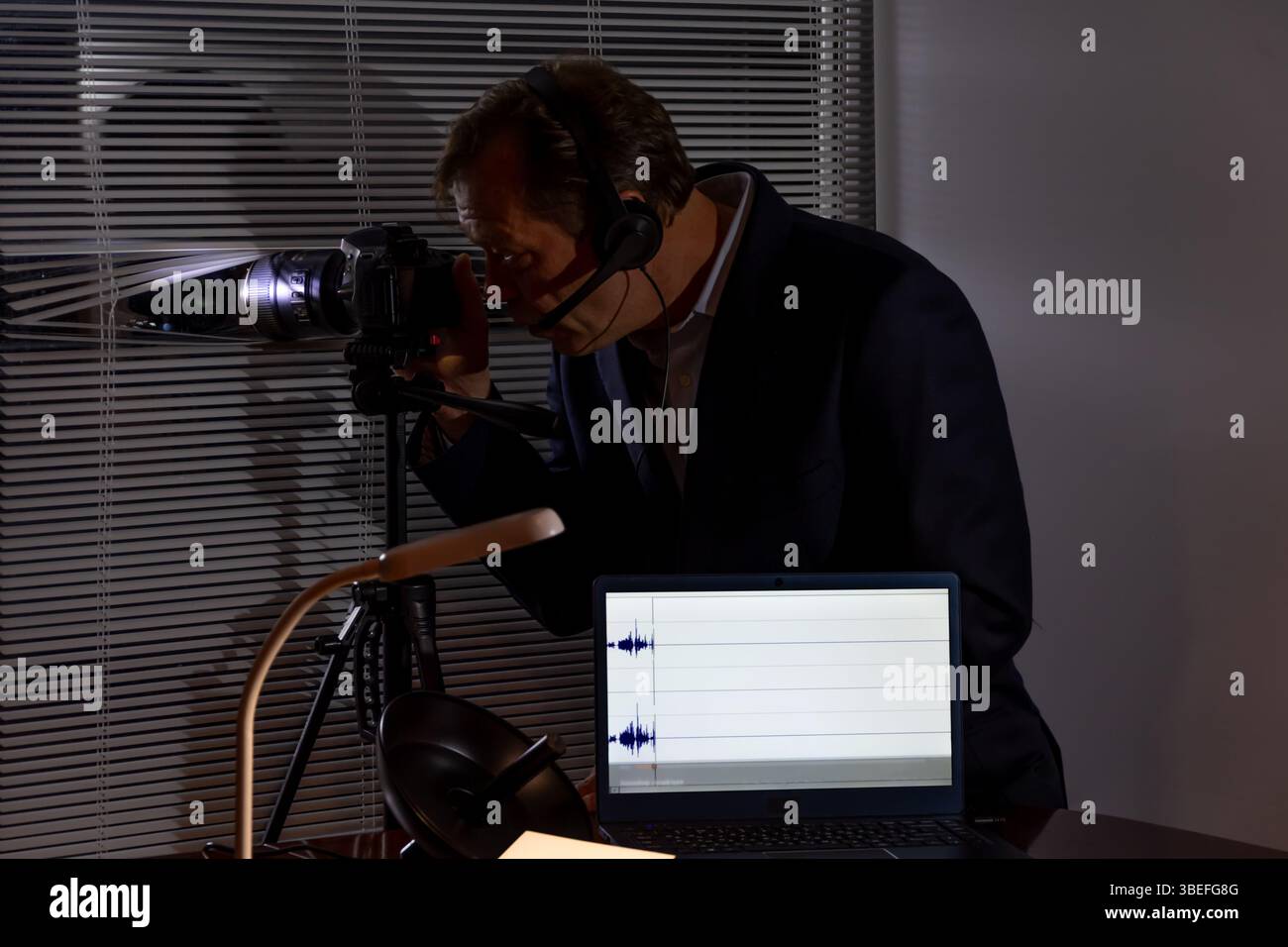 A private detective in a dark room with a camera and laptop records ...