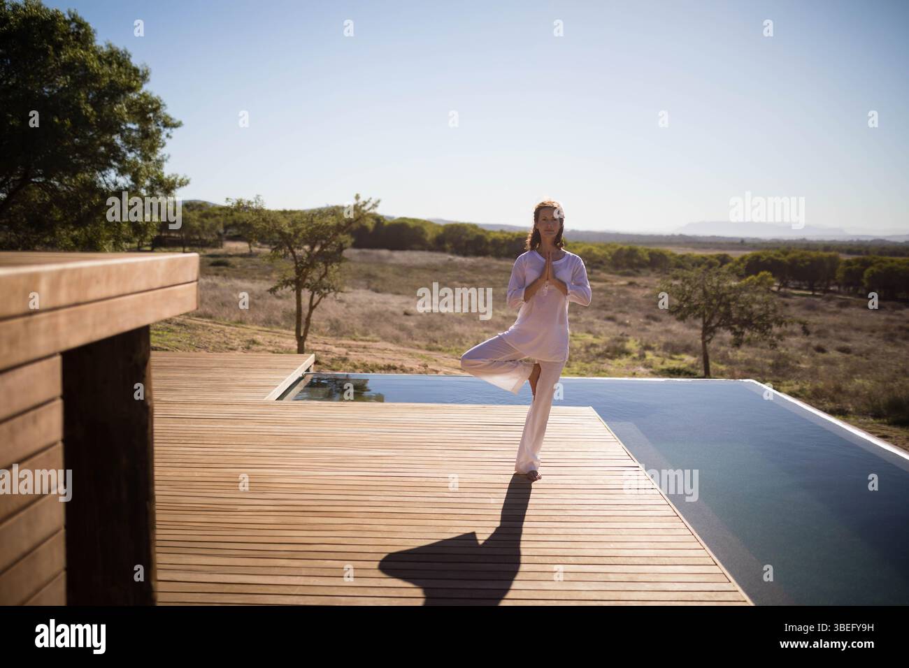 woman practicing yoga in tree pose on villa terrace beside infinity ...