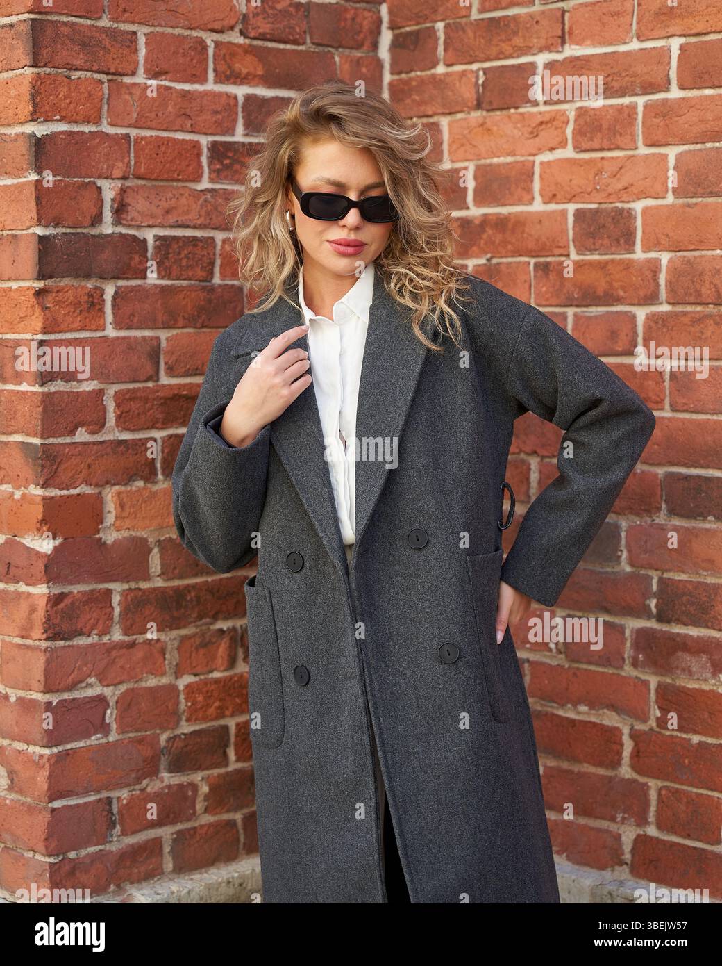 Blonde woman in stylish coat poses against red brick wall on a sunny ...