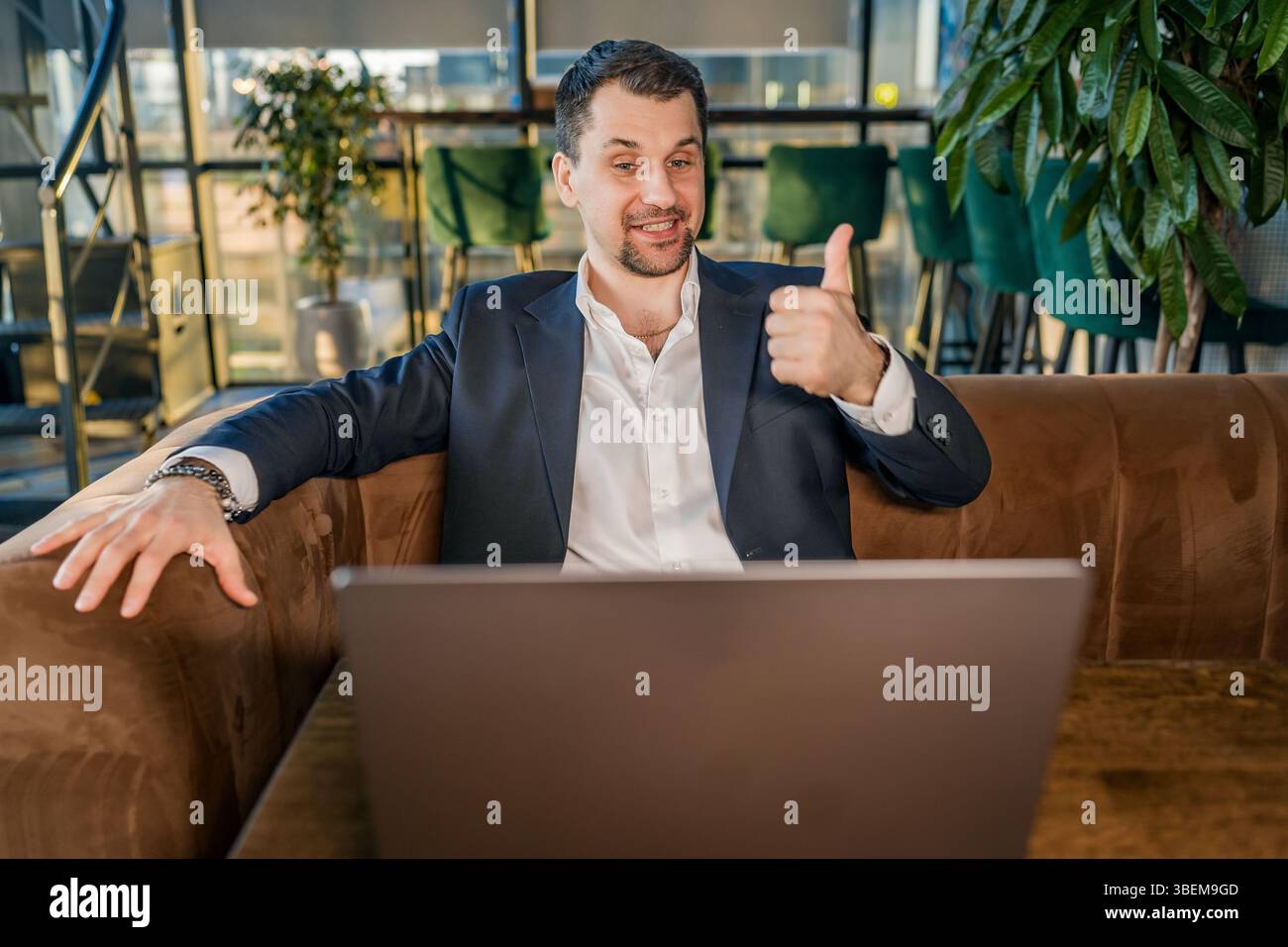 A smiling businessman sits with a laptop in a cozy cafe, giving a ...
