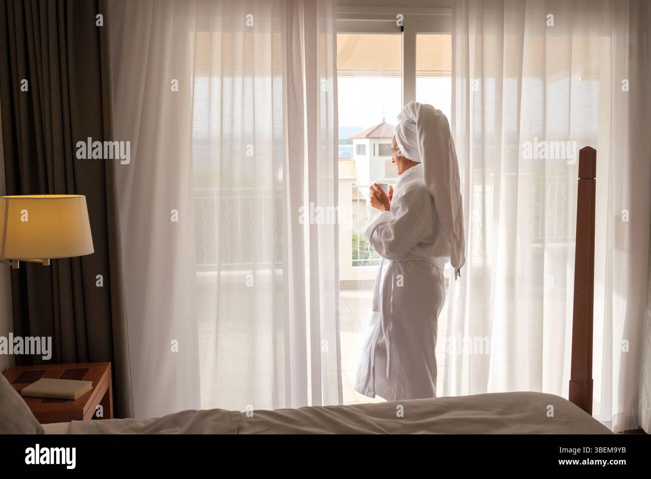 A person in a white robe stands by a large window with sheer curtains ...