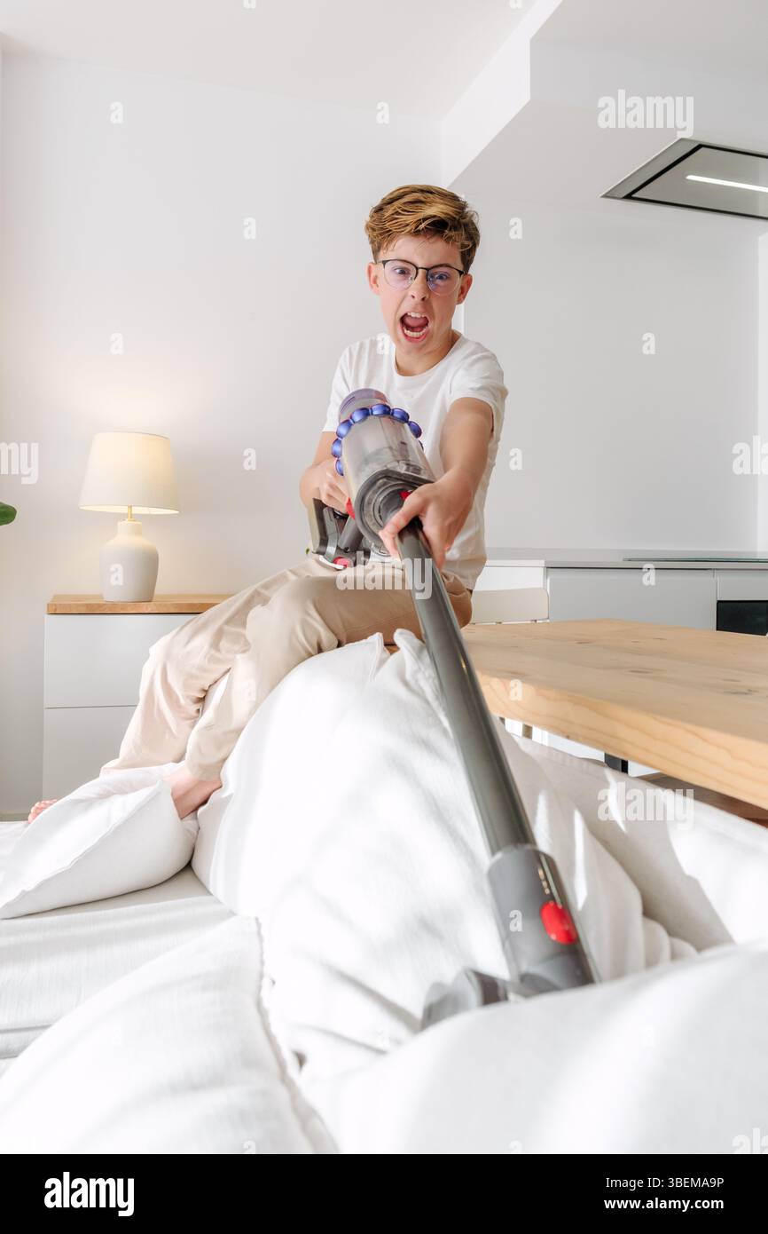 A boy vigorously using a vacuum cleaner on a sofa in a bright modern ...