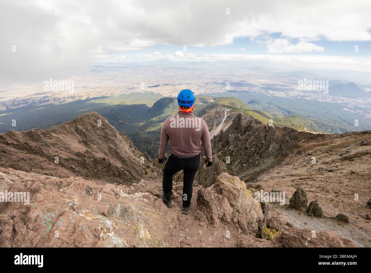 Hiker in helmet stands on a rocky peak, overlooking expansive valleys ...
