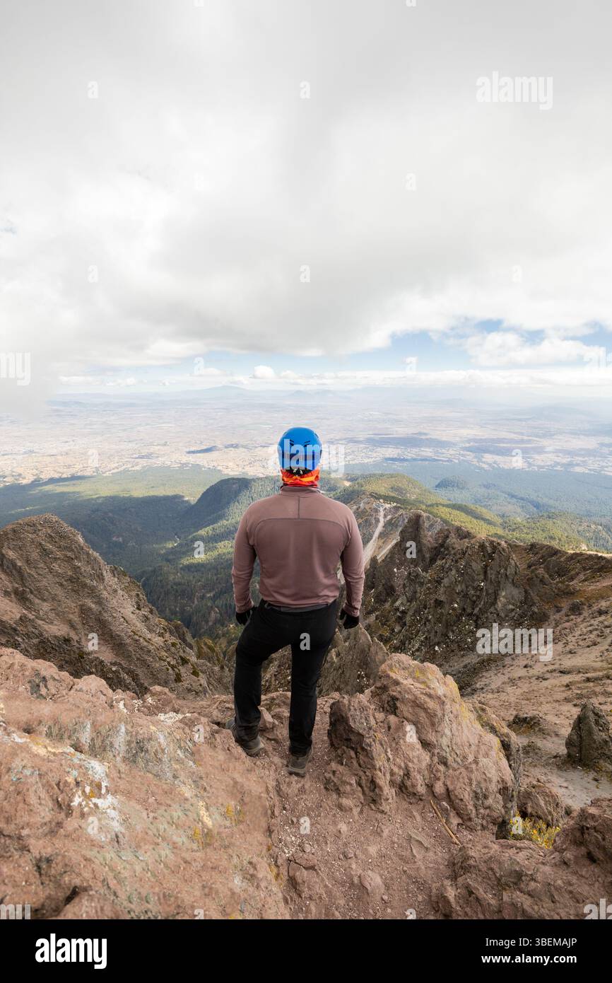 A lone hiker stands on a rocky ledge, overlooking expansive mountainous ...
