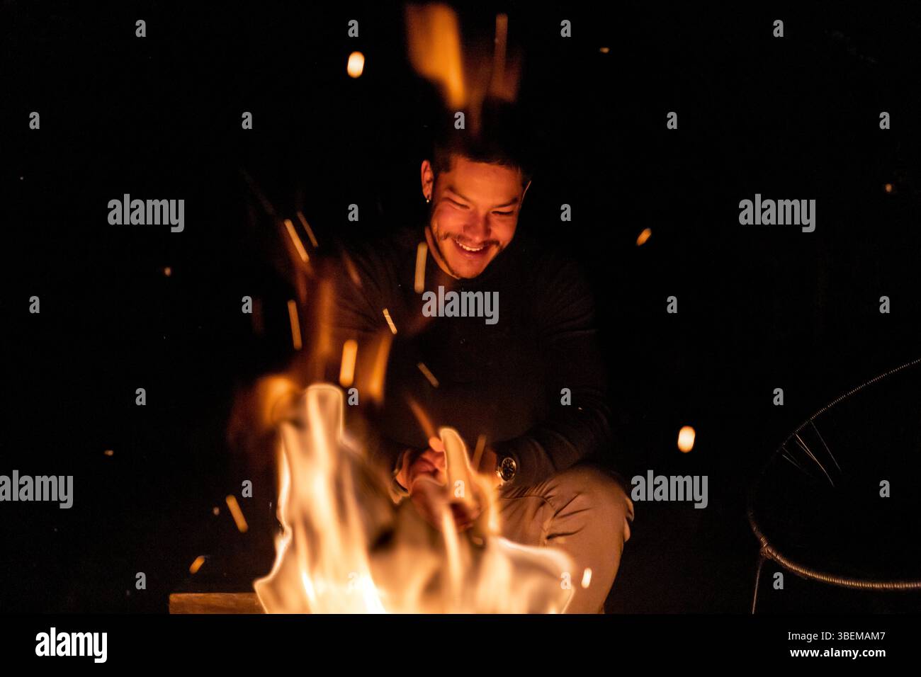 A man sits by a campfire, illuminated by the warm glow of the flames in ...
