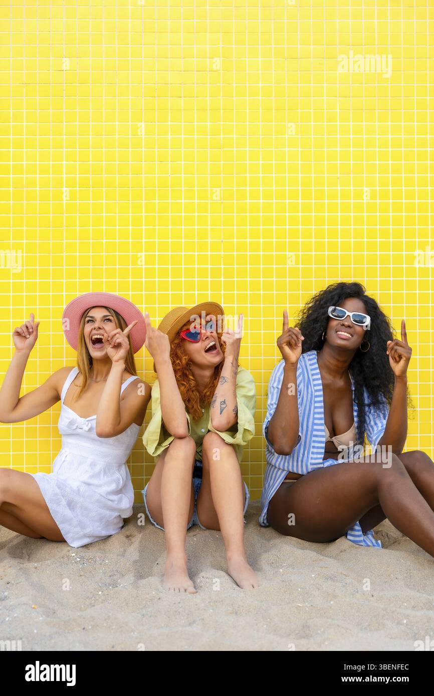 Three diverse women wearing summer clothes and sunglasses are sitting on the sand, pointing ...