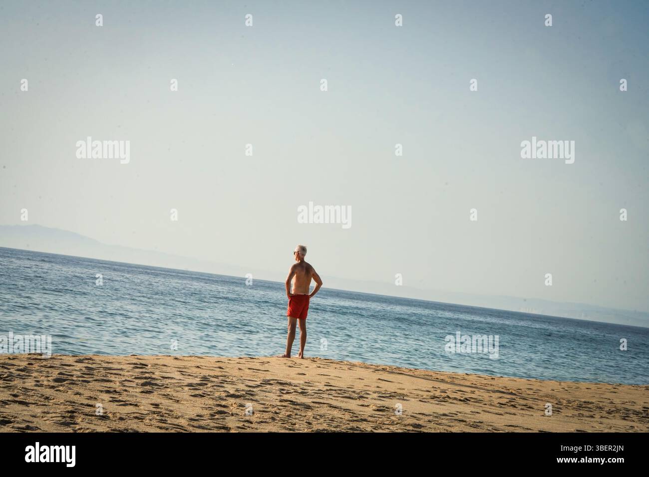 Senior man standing alone on a quiet beach, gazing at the sea—capturing ...