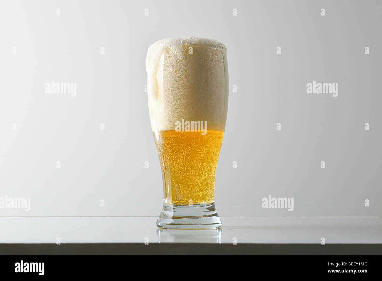 Glass of beer with lots of foam overflowing, reflected on a white table ...