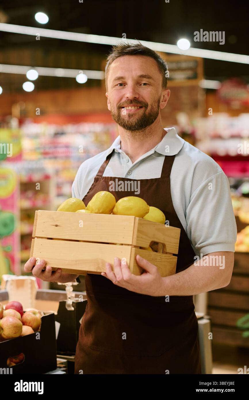 Smiling grocer wearing apron, holding wooden crate of fresh mangoes in ...