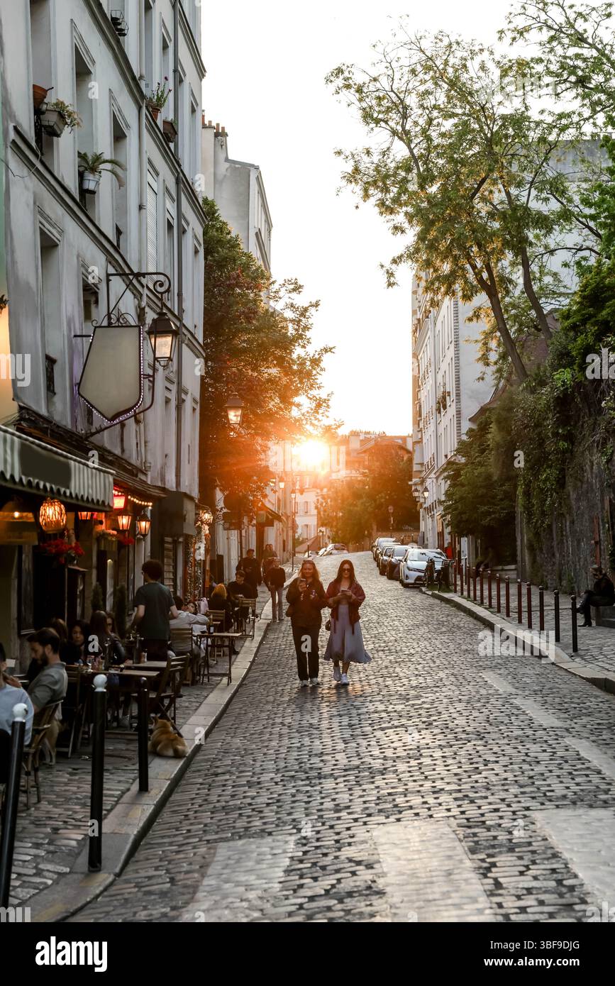 The setting sun casts a warm, golden flare down a lively cobblestone ...