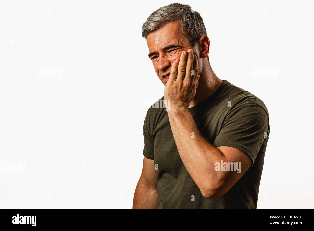 A man stands in a studio, looking pained as he touches his cheek ...